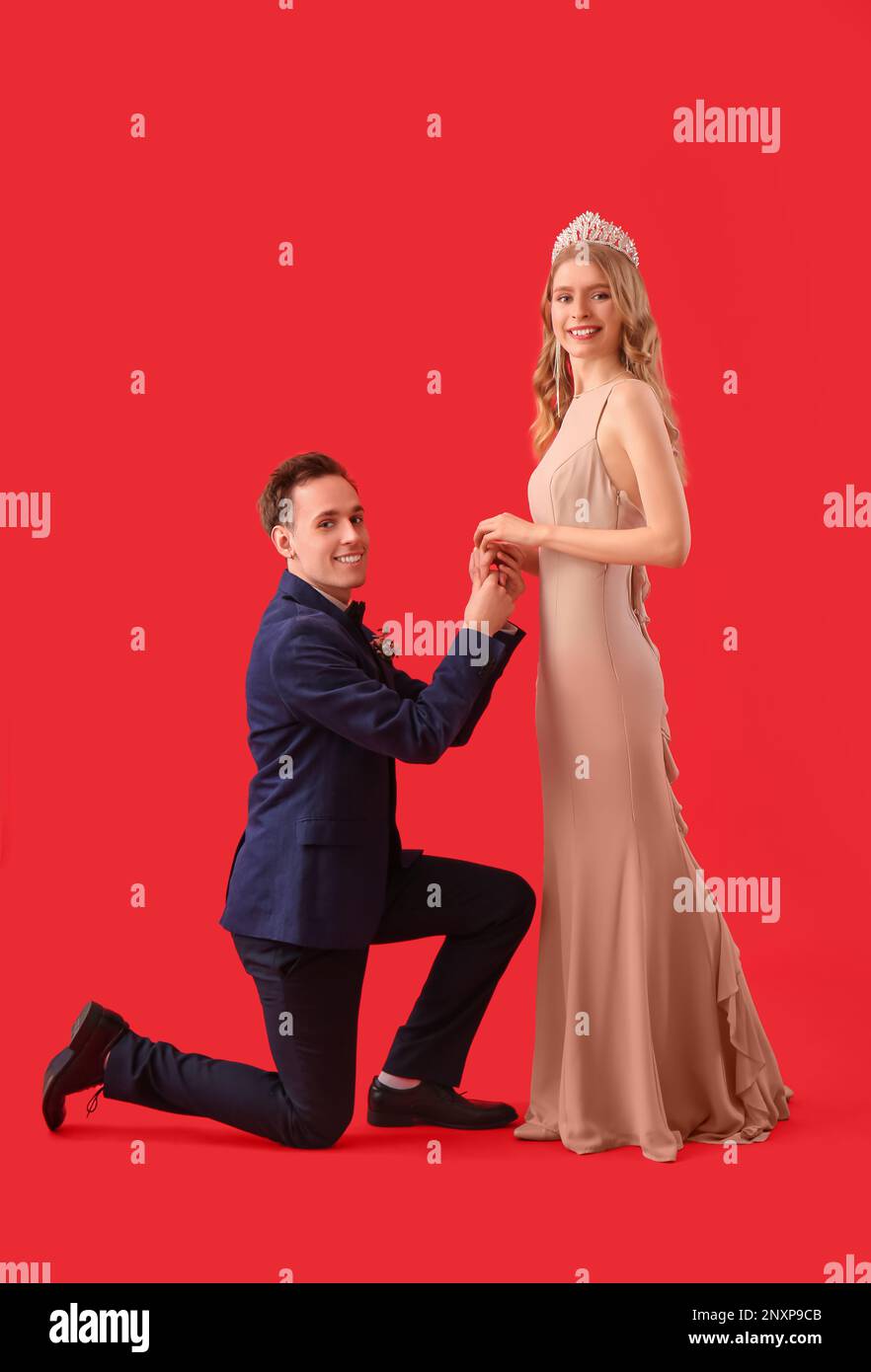 Young man standing on one knee in front of his prom date against red ...