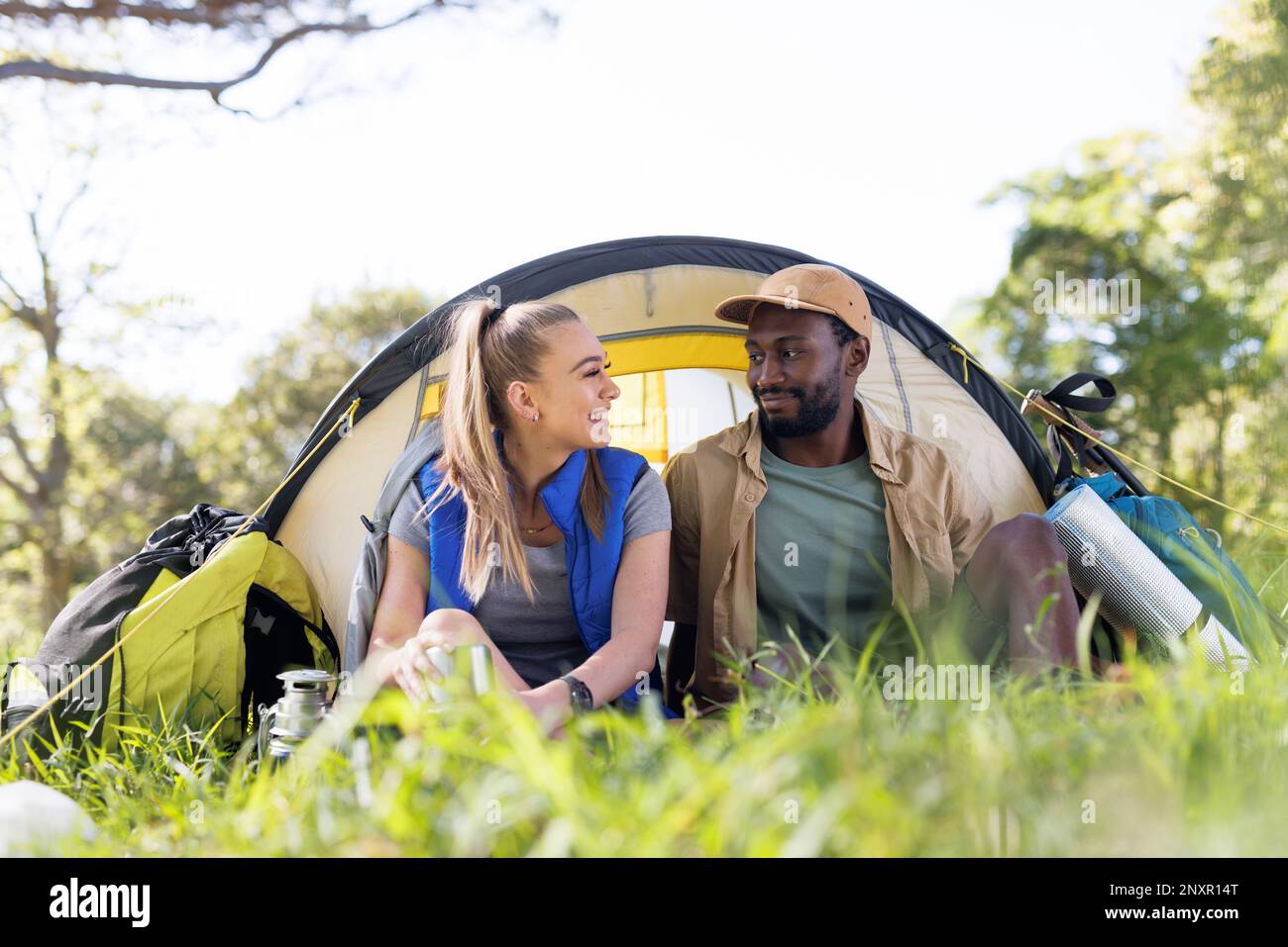Happy, diverse couple camping in forest sitting in front of tent ...