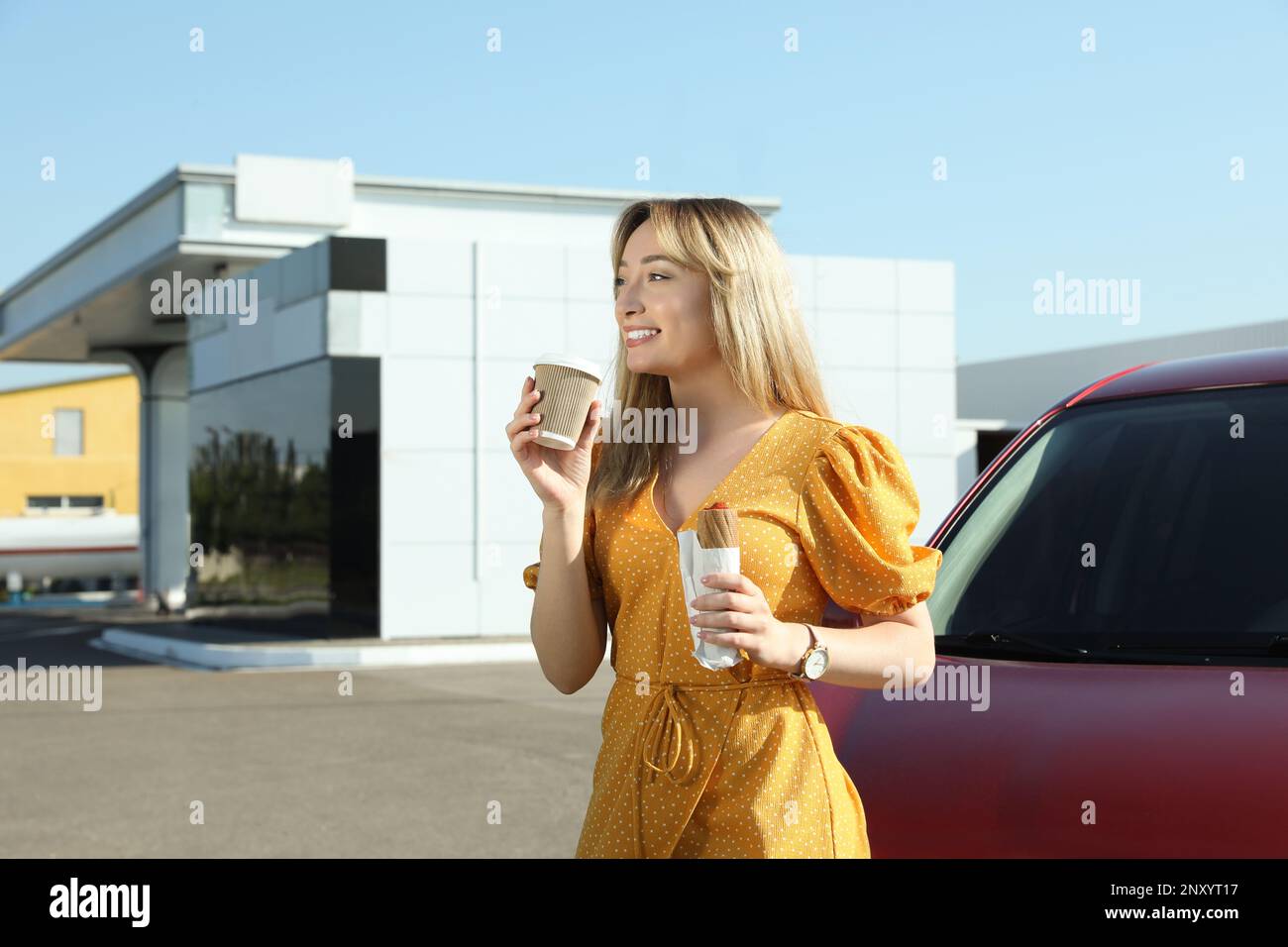 Beautiful young woman with hot dog drinking coffee near car at gas ...