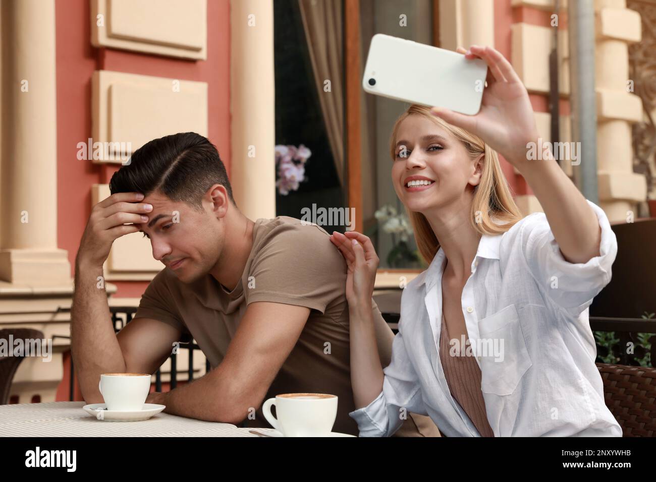 Young woman trying to take selfie with her displeased boyfriend in outdoor cafe. Boring date ...