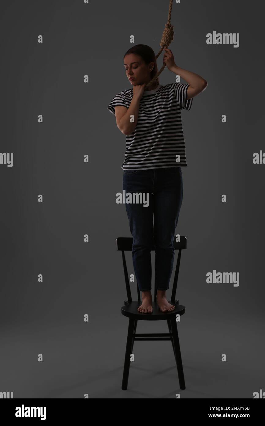 Depressed woman with rope noose standing on chair against grey ...