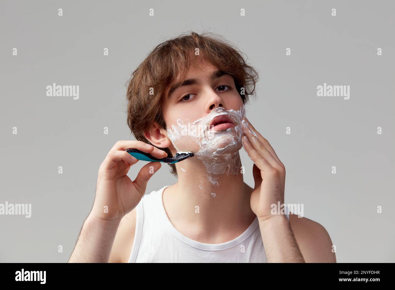 Portrait of young man shaving face with razor and cream, posing against ...
