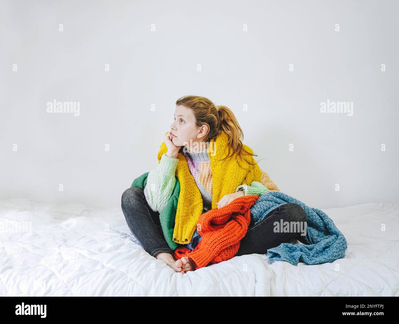 Full body of thoughtful young female sitting on comfortable bed in pile ...