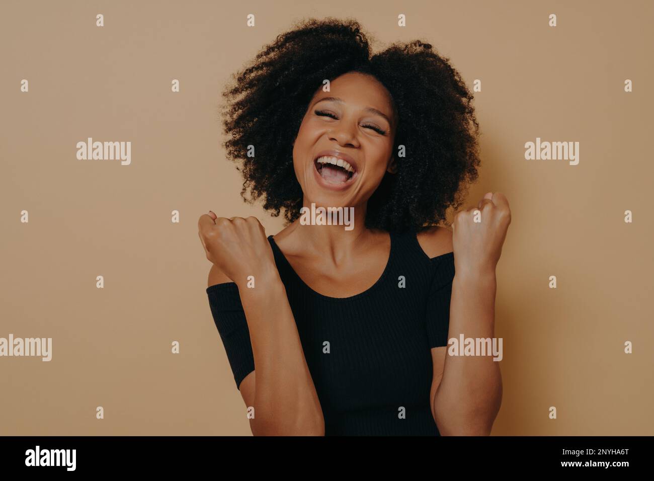 Studio shot of young joyful excited dark skinned woman keeping fists ...
