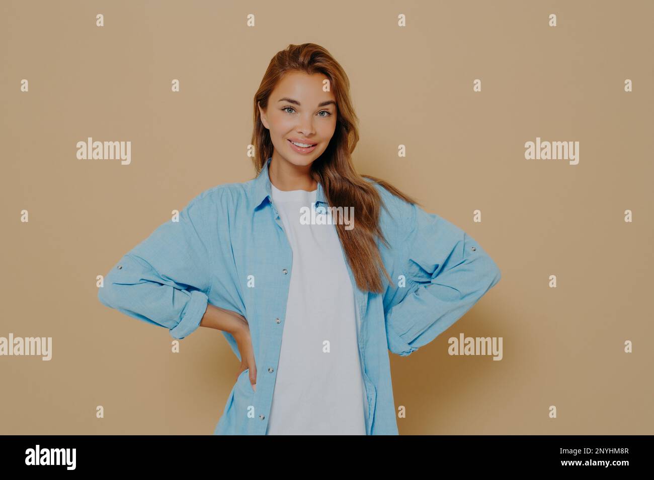 Self assured smiling lovely model keeps hands on waist, wears long blue ...