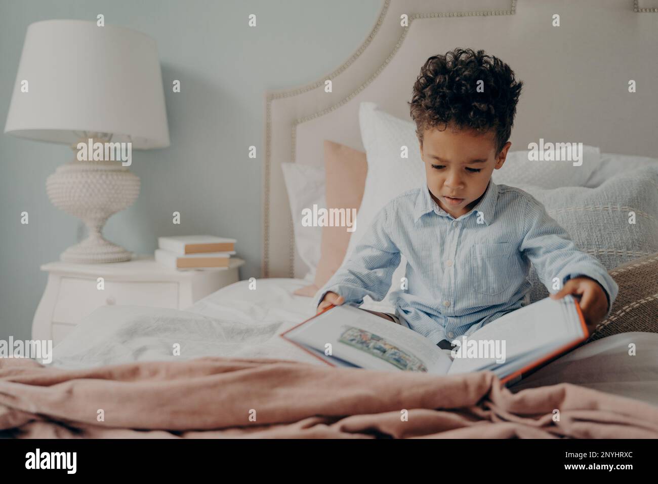 Cute focused small boy in casual wear holds and tries to read childrens ...