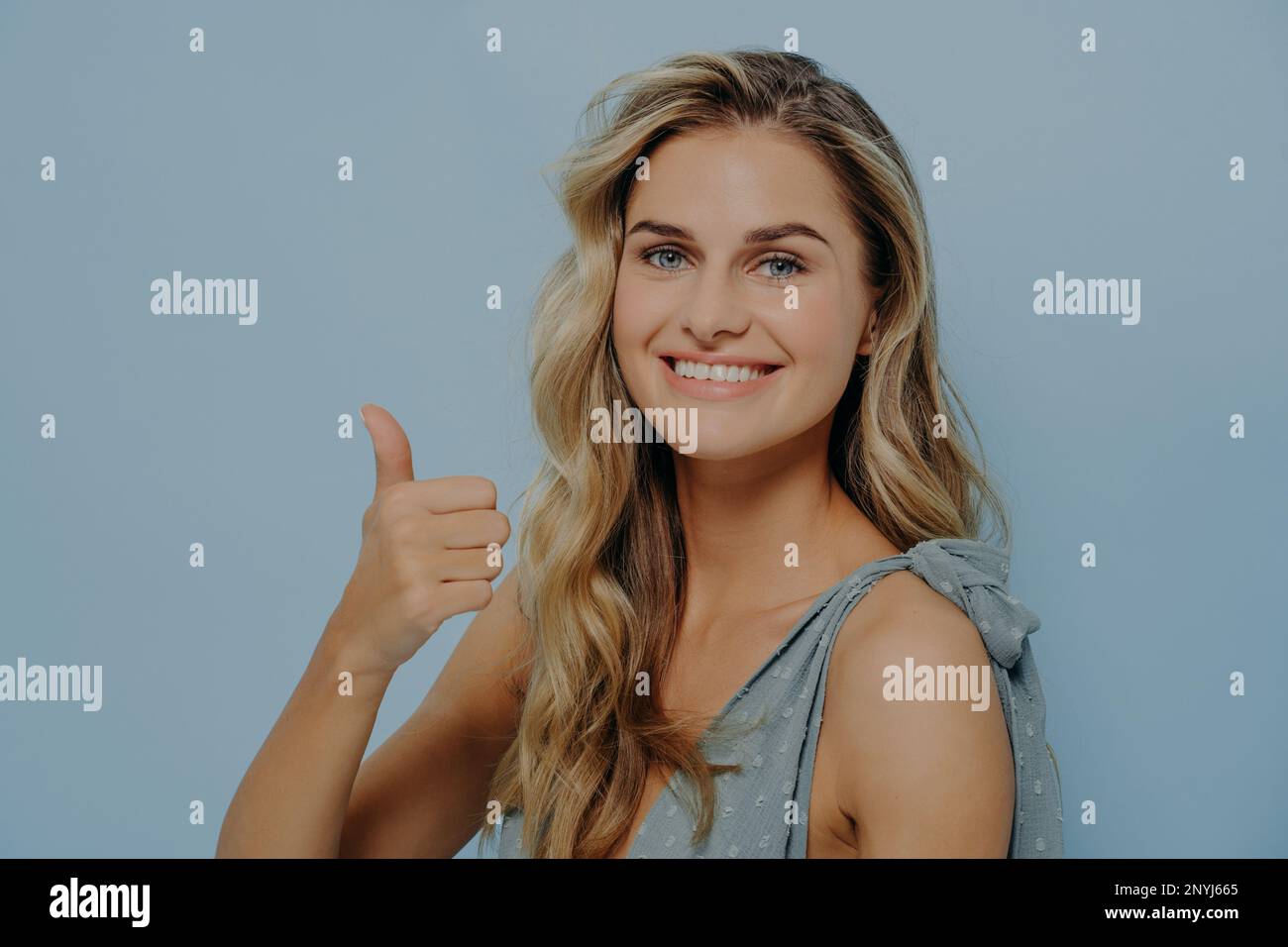 Cheerful blonde woman showing thumbs up gesture while standing slightly ...