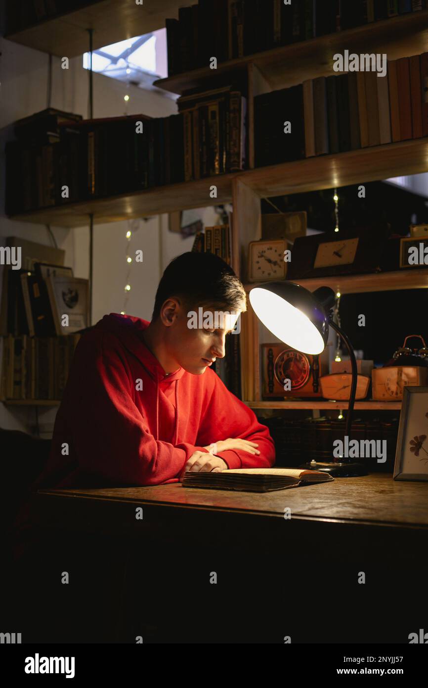 Young man is reading an old book in the library under a table lamp ...