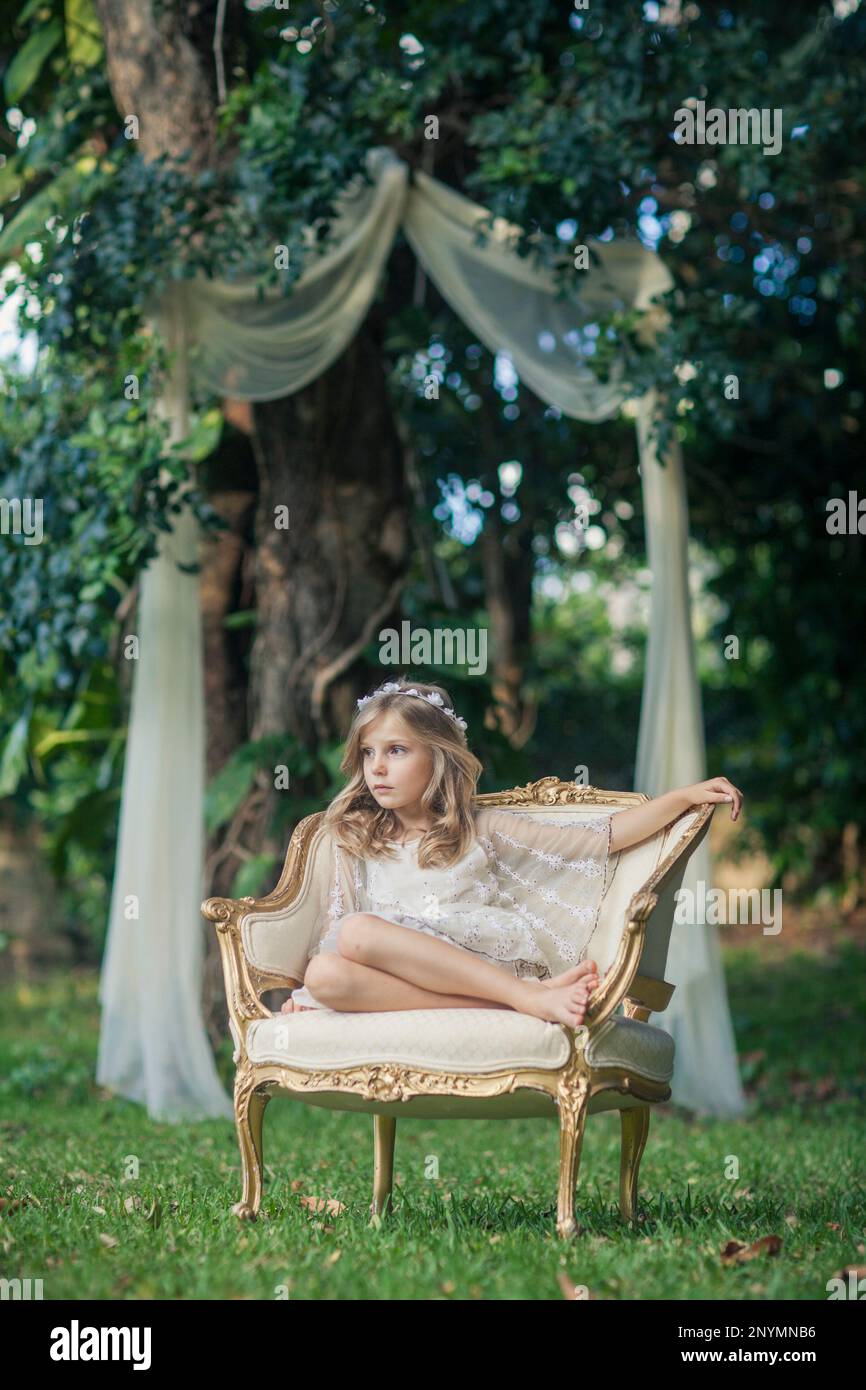 Little girl barefoot in white dress and flower crown sitting on antique ...