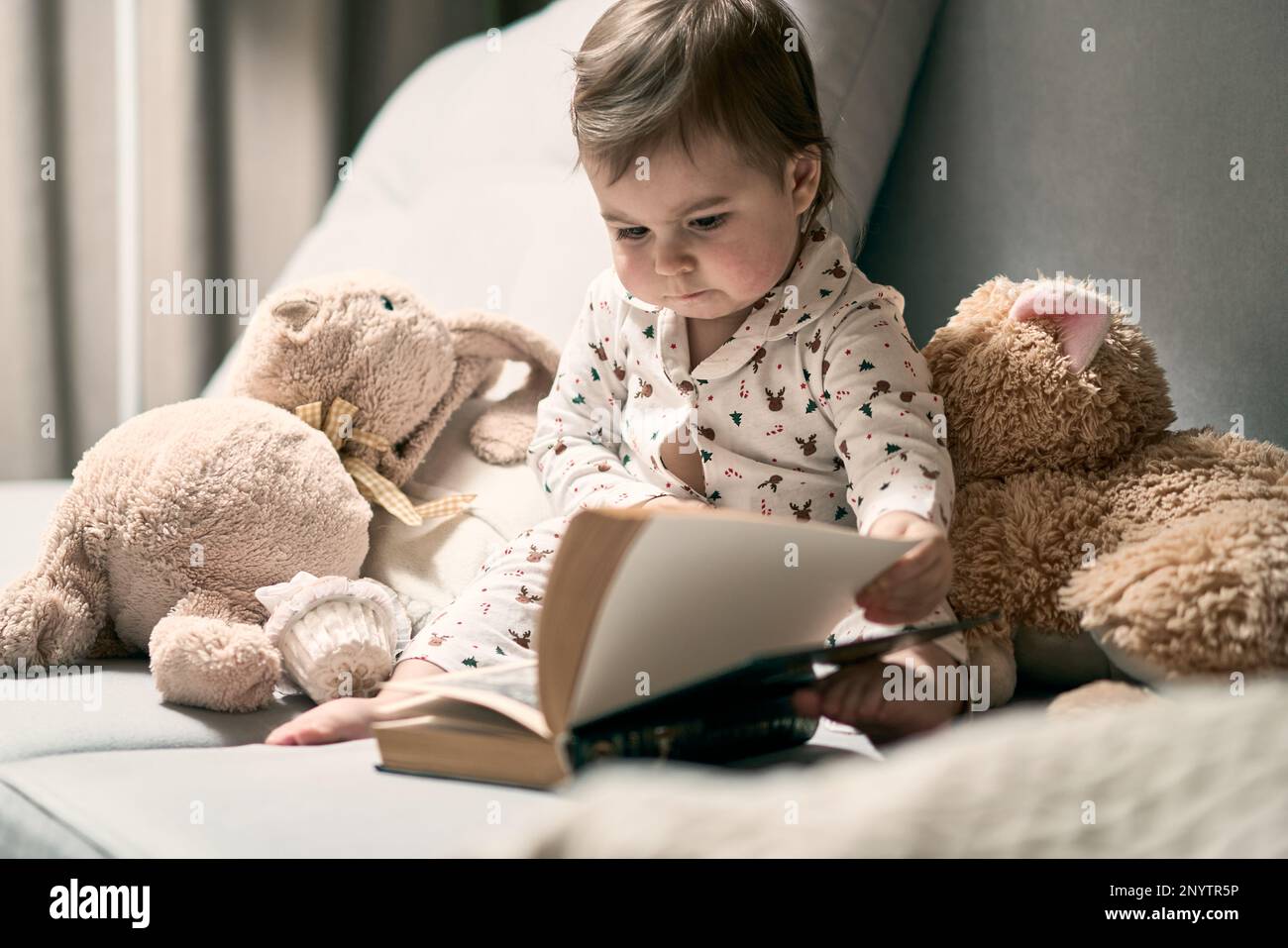 A little baby sitting on the sofa reading book. Teddy bear and rabbit ...