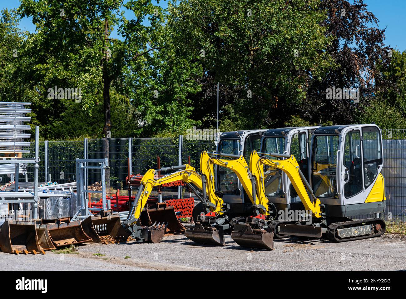 Excavators standing in a row. Metal fence and trees in the background ...