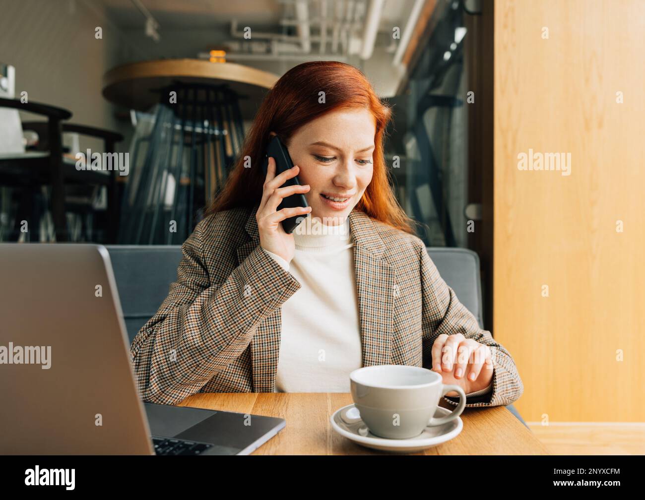 Young woman with ginger hair in formal wear talking on a mobile phone while sitting at a cafe ...