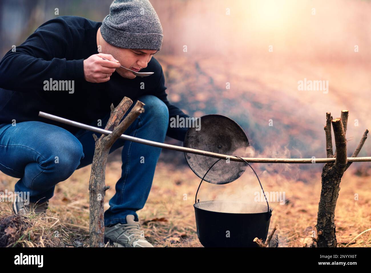A young man cooks food on an open fire at sunset and tastes the broth ...