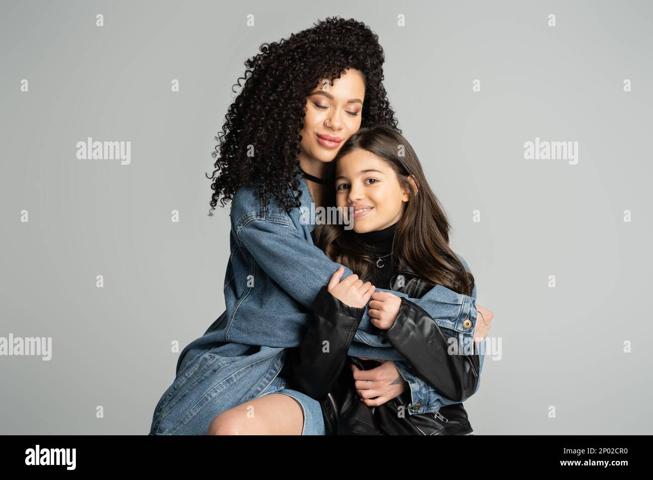 Curly woman in denim jacket hugging happy daughter isolated on grey ...