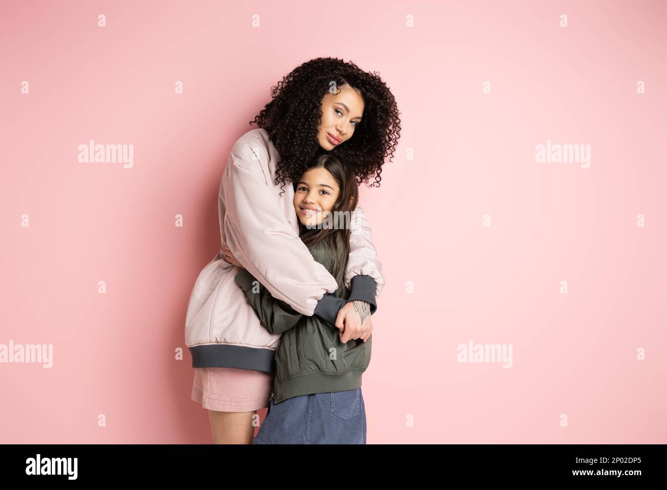 Curly woman in bomber jacket hugging daughter on pink background,stock ...