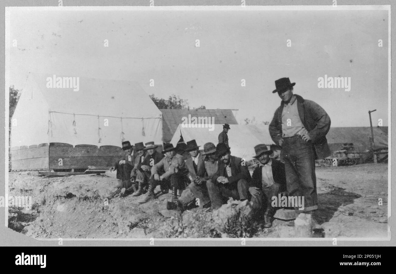 Men sitting along gulch; tented structures in background. Frank and ...