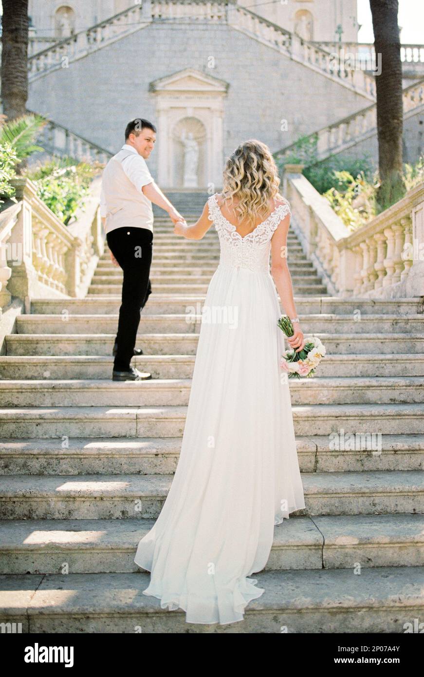 Bride follows groom holding his hand on the stone steps. Back view ...