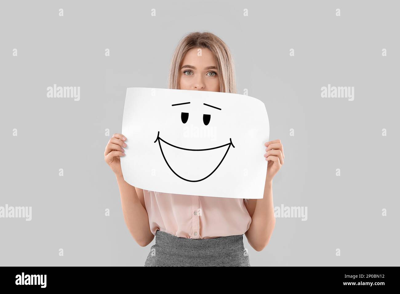 Pretty young woman holding paper with drawn smiling emoticon on light ...