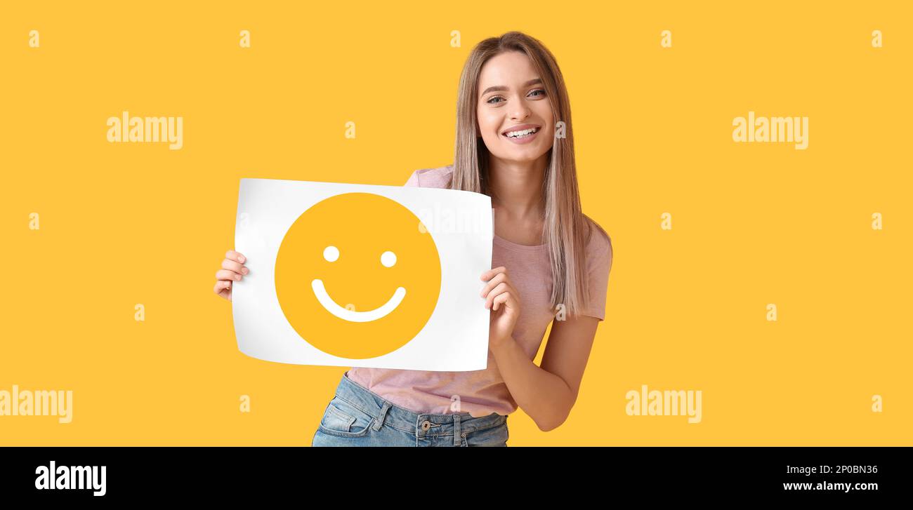 Pretty young woman holding paper with smiling emoticon on yellow ...