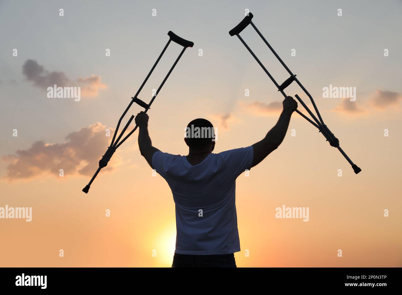 Man raising hands with underarm crutches up to sky outdoors at sunset ...