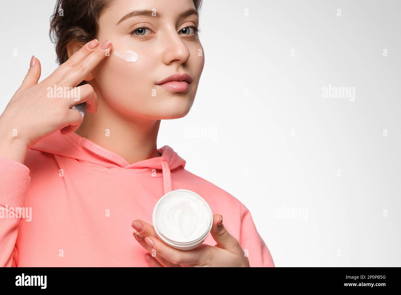 A young girl with blue eyes applies cream to her face and holds a jar of thick facial ...