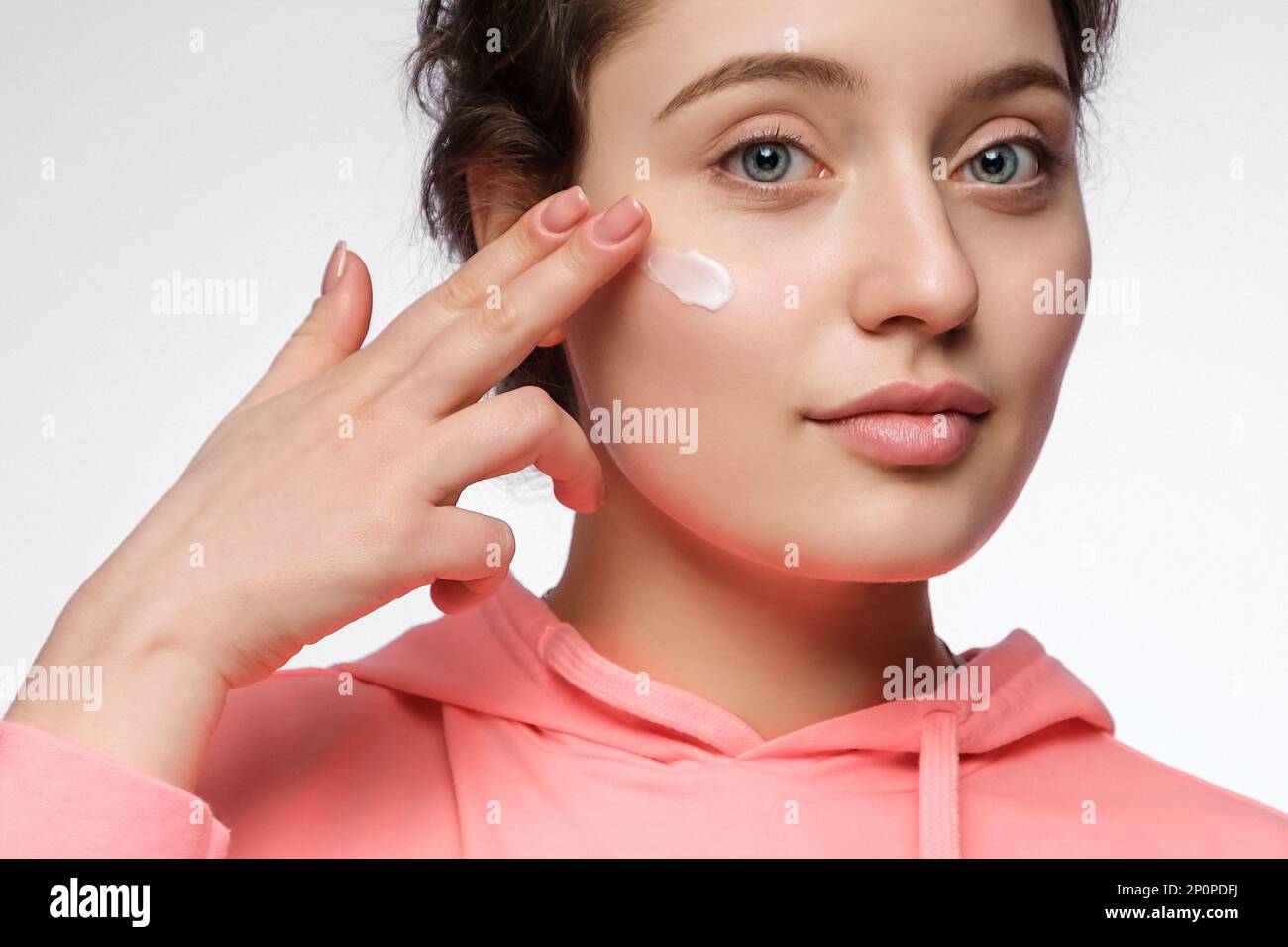 A young girl with blue eyes applies cream to her face and holds a jar ...