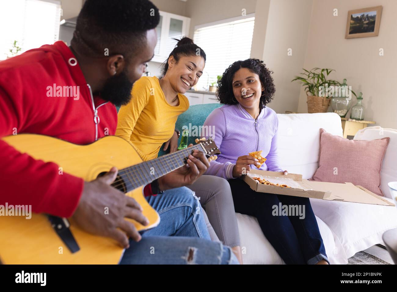 Happy diverse friends sitting on sofa, playing guitar and eating pizza ...