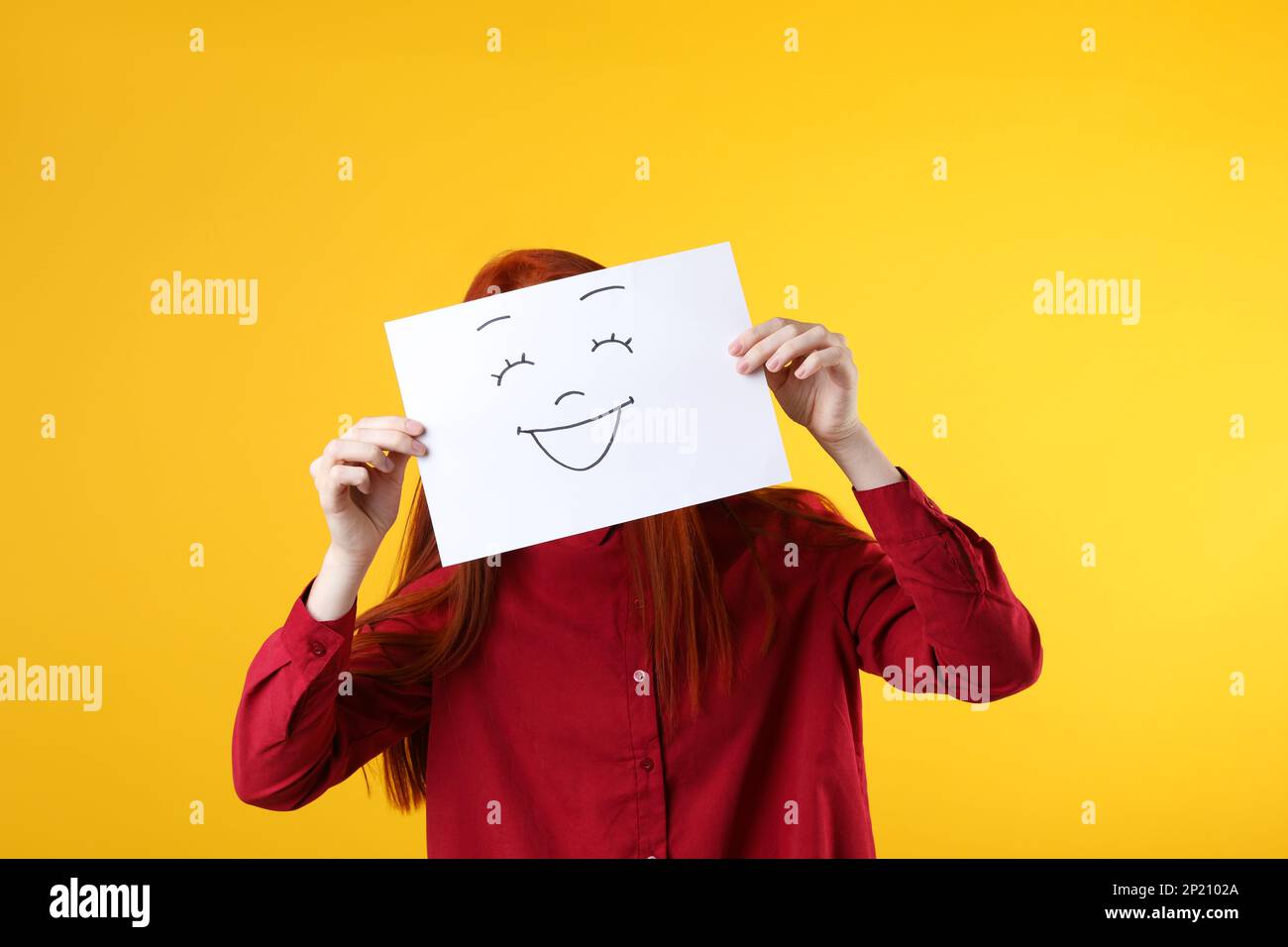 Woman hiding behind sheet of paper with happy face on yellow background ...
