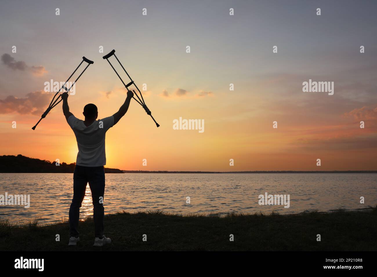 Man raising hands with underarm crutches up to sky near river at sunset ...