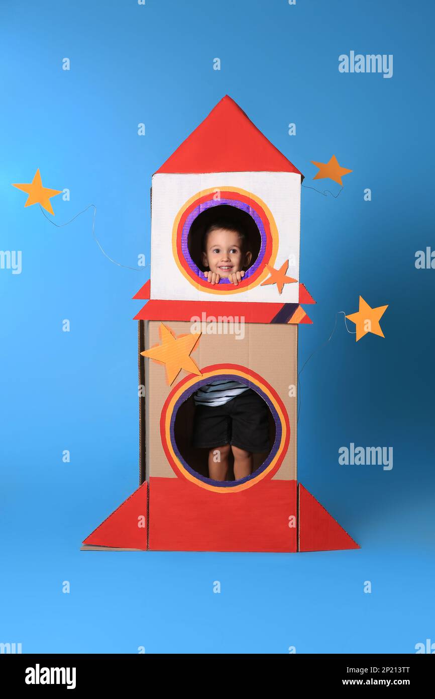 Cute little boy playing with cardboard rocket on light blue background ...