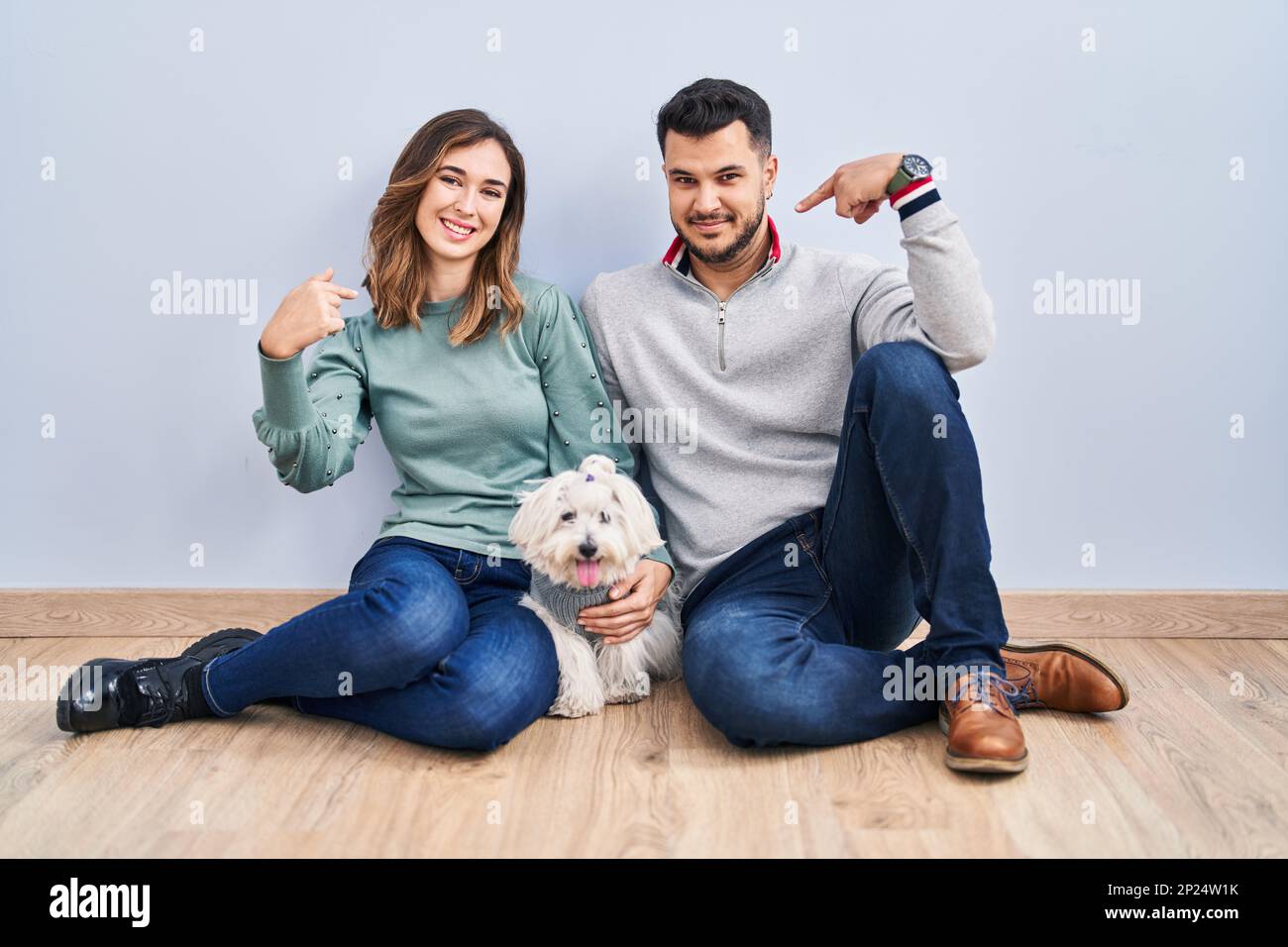 Young hispanic couple sitting on the floor with dog looking confident ...