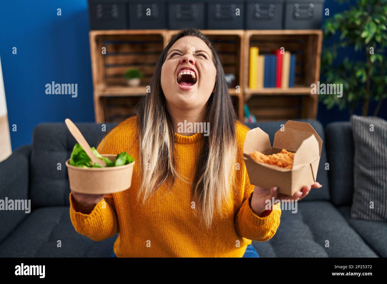 Young hispanic woman holding healthy salad and fried chicken wings ...