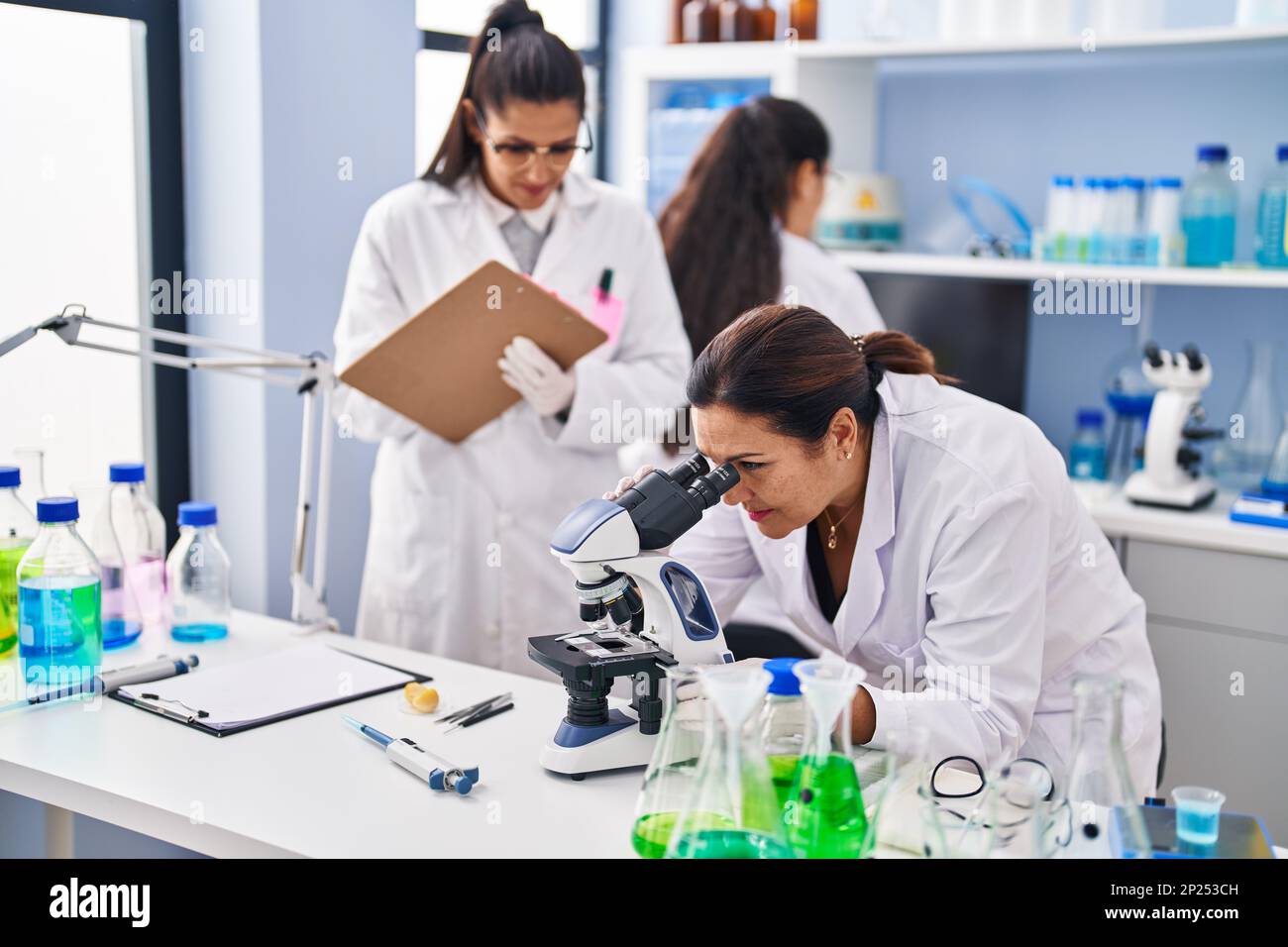 Three woman scientists using microscope write on checklist at ...