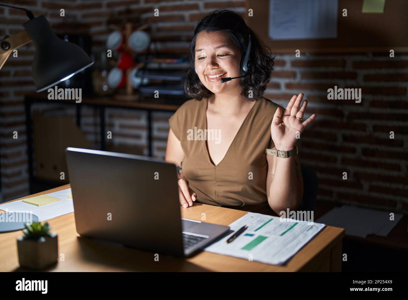 Young hispanic woman working at the office at night waiving saying ...