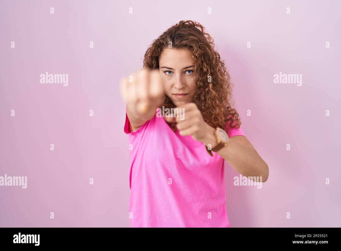 Young caucasian woman standing over pink background punching fist to ...