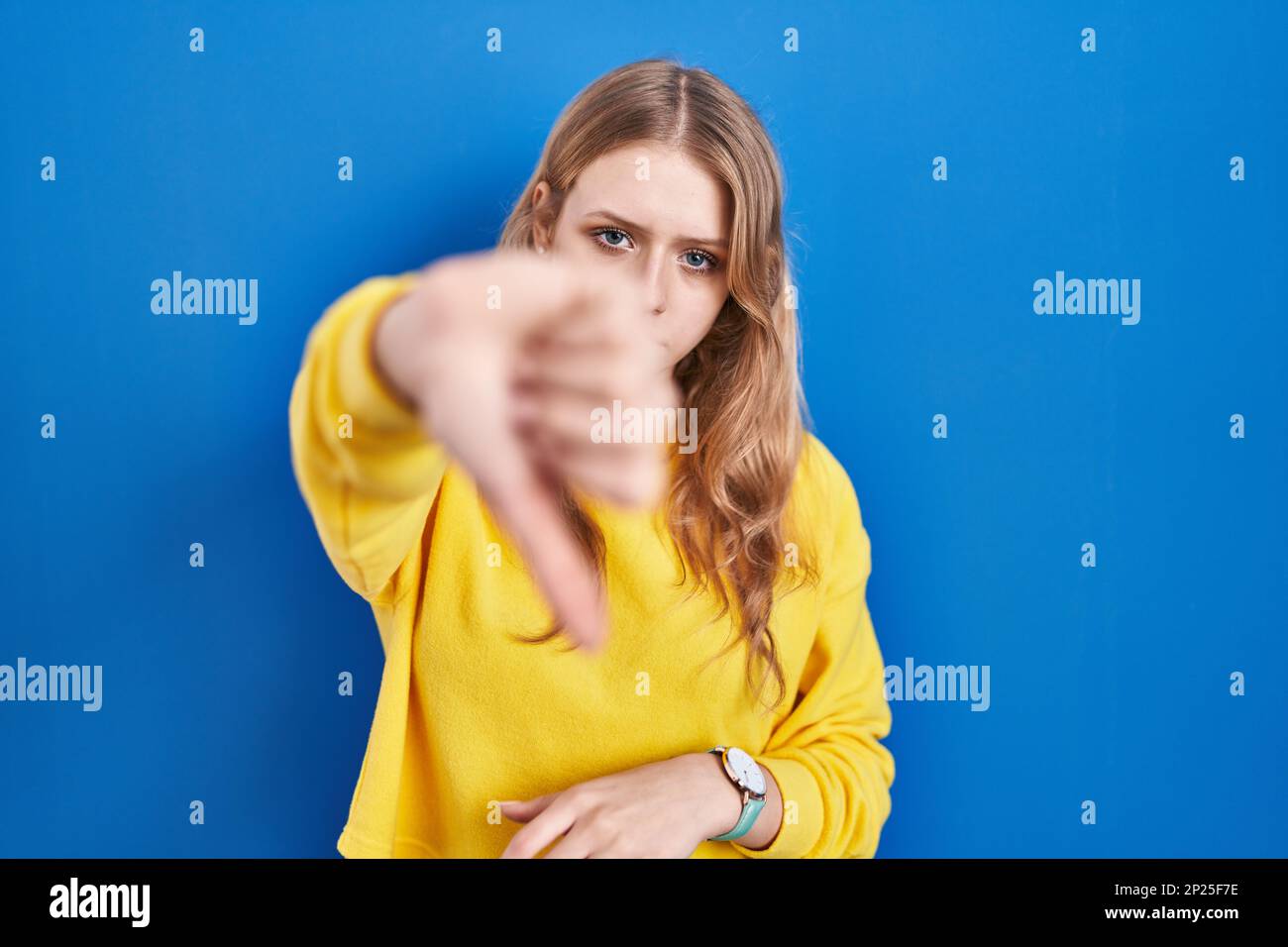 Young caucasian woman standing over blue background looking unhappy and ...