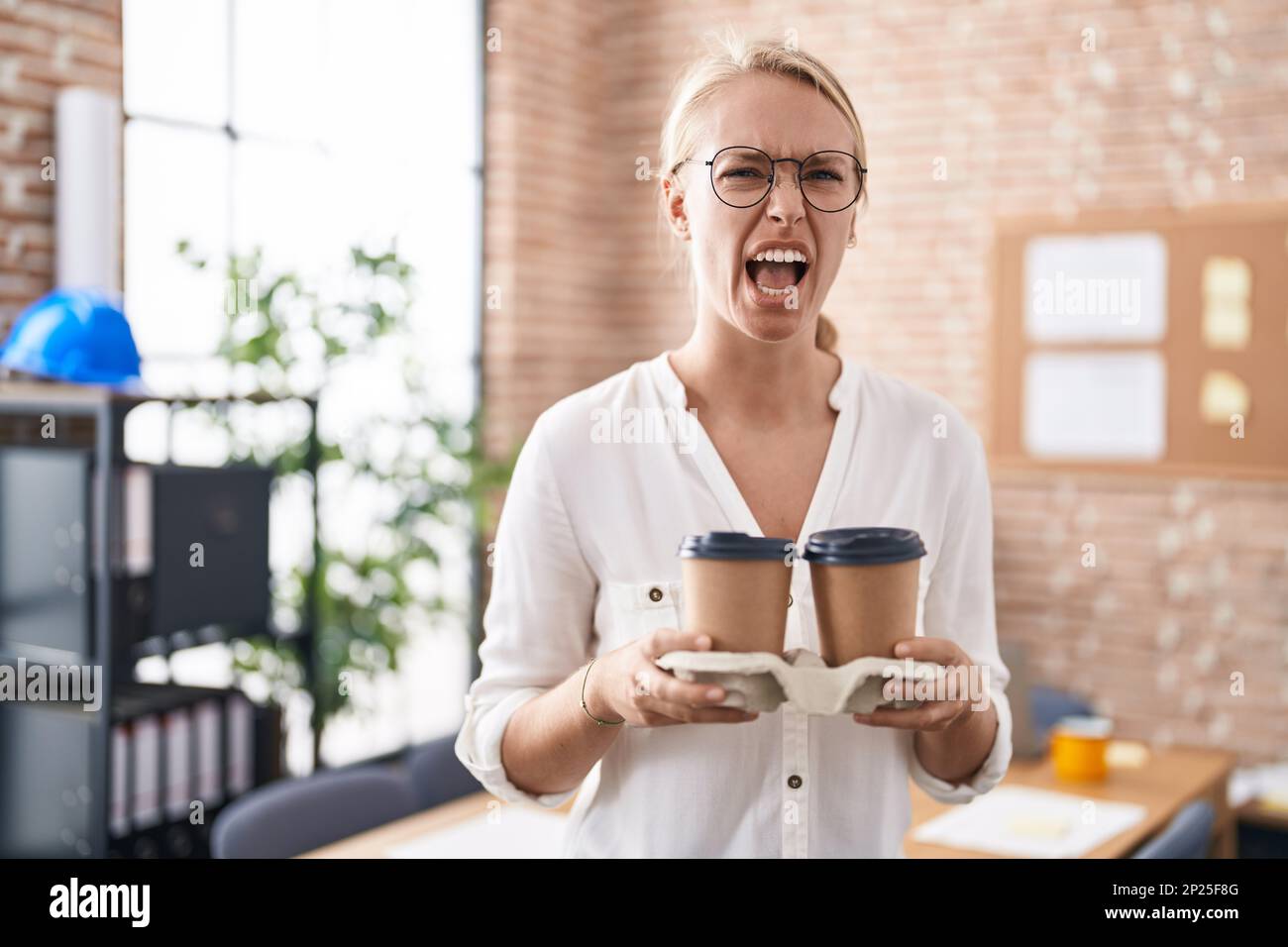 Young caucasian woman working at the office holding coffee cups angry ...