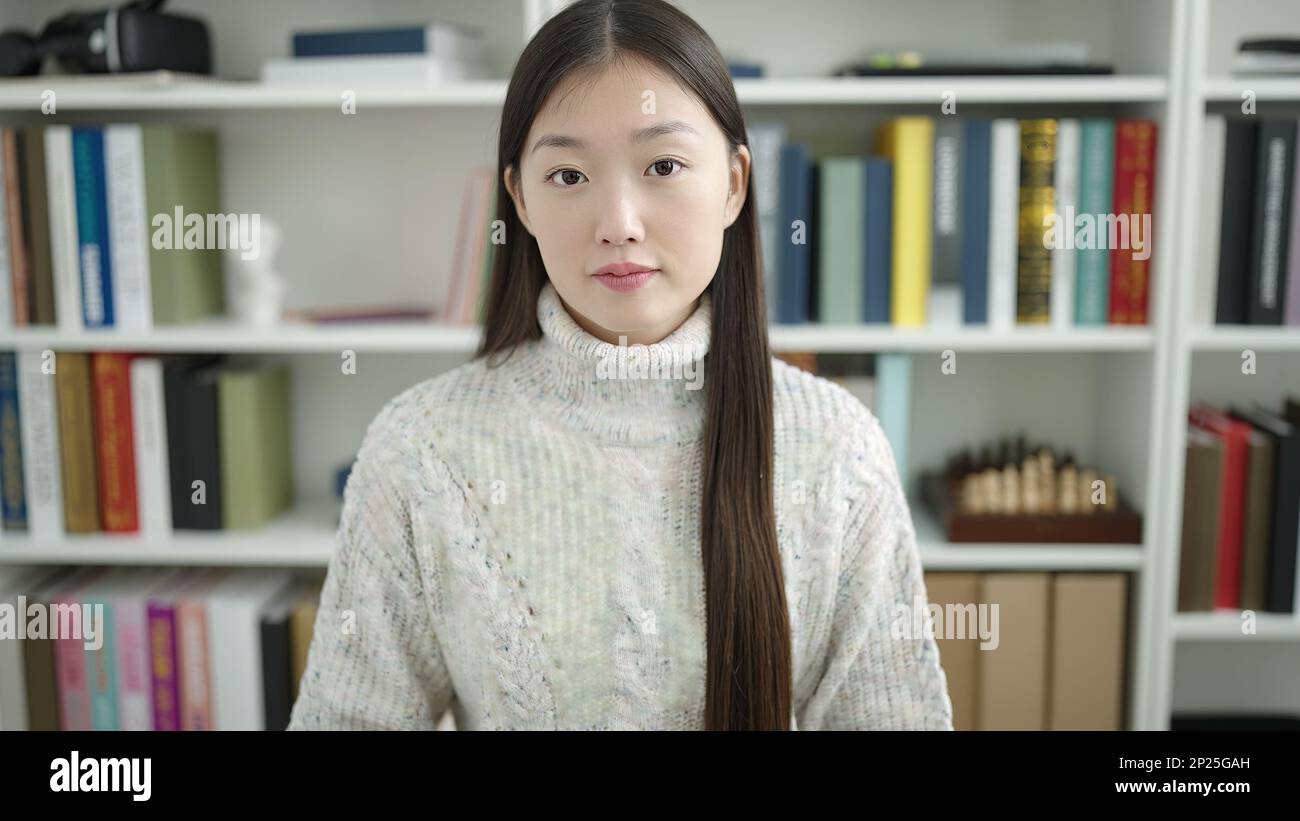 Young chinese woman student standing with relaxed expression at library ...