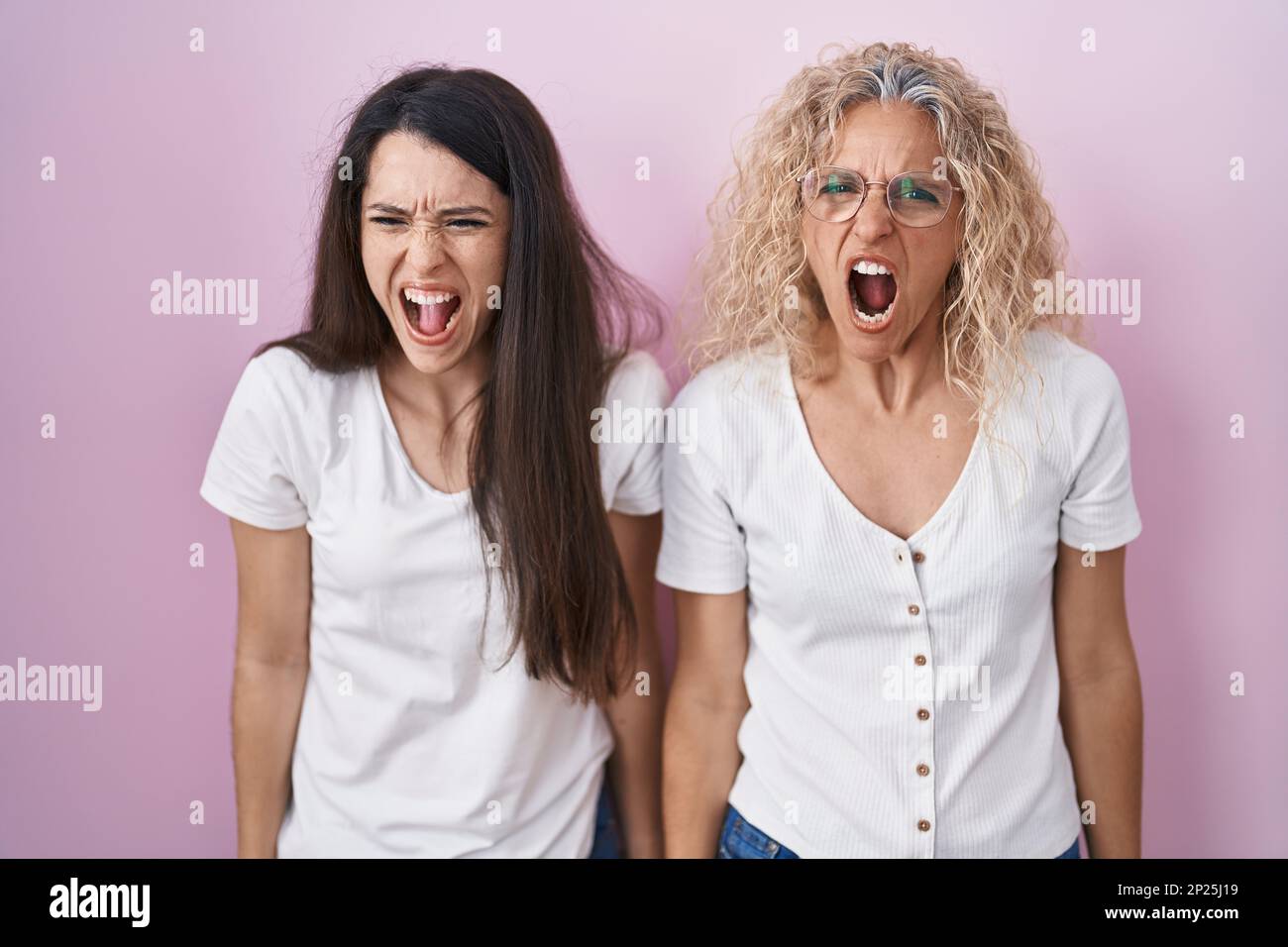 Mother and daughter standing together over pink background angry and ...