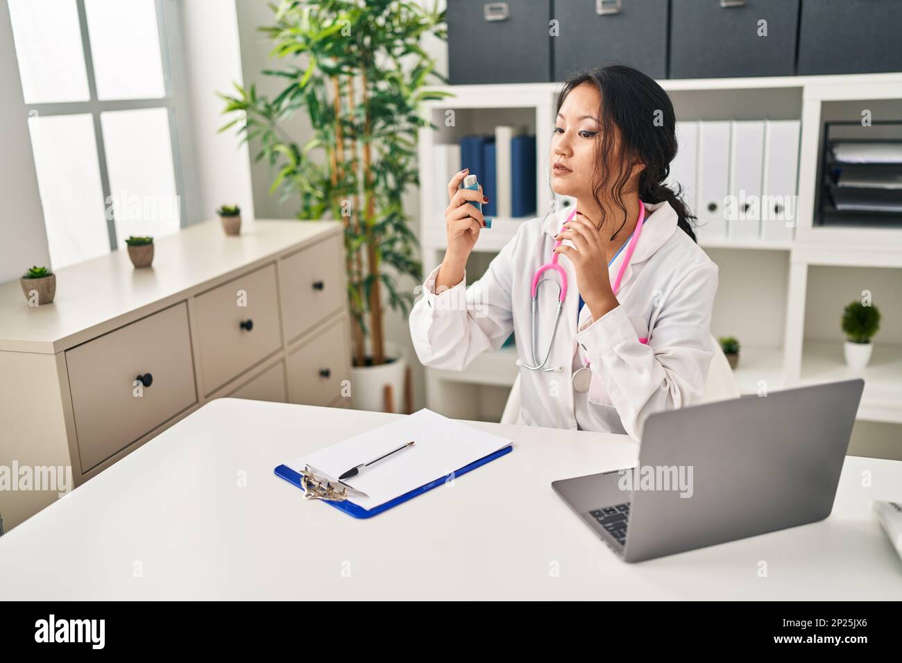 Young chinese woman wearing doctor uniform holding inhaler at clinic ...