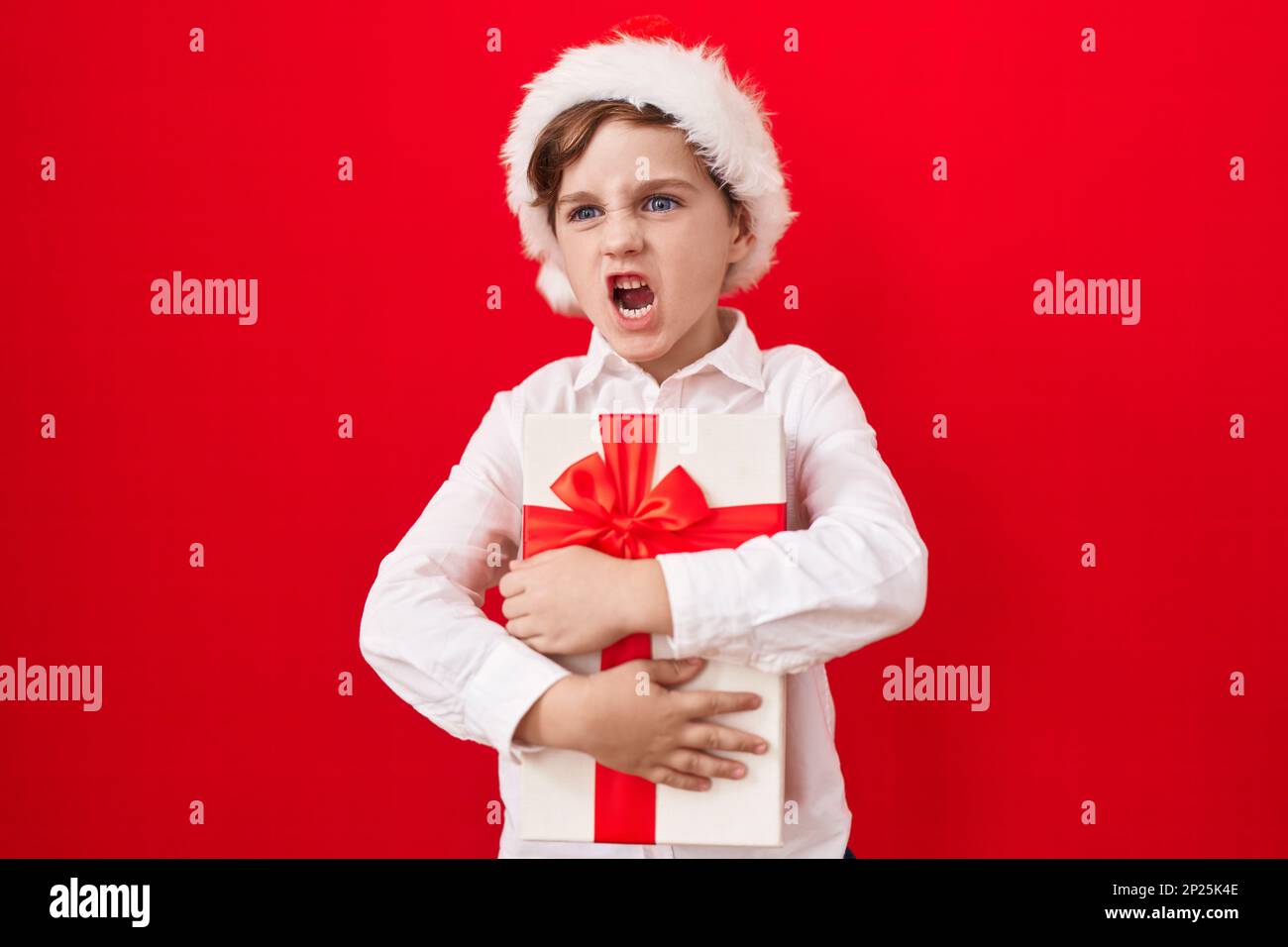 Little caucasian boy wearing christmas hat and holding gifts angry and ...