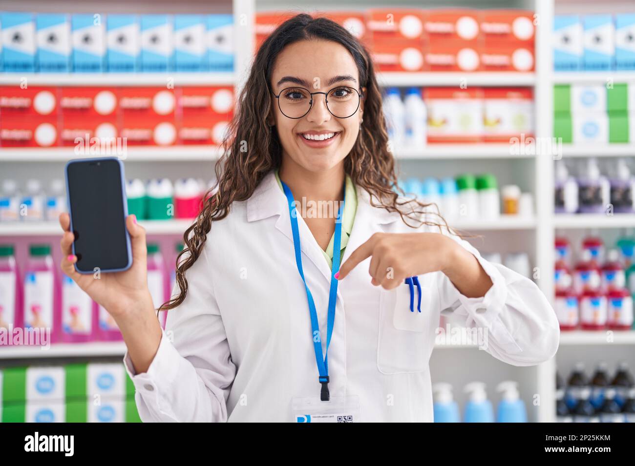 Young hispanic woman working at pharmacy drugstore showing smartphone ...