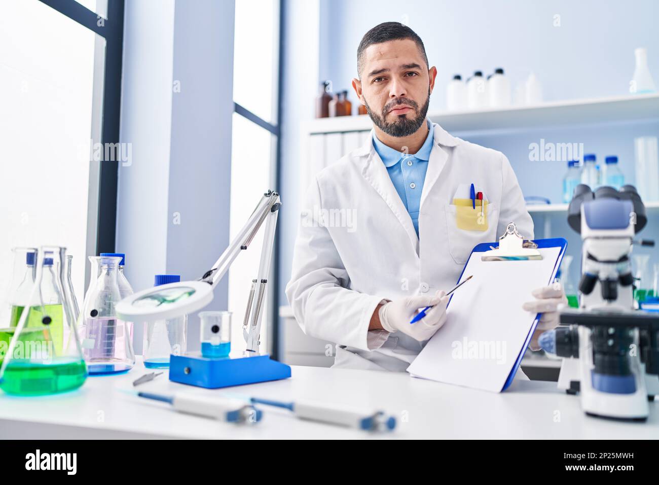Hispanic man working at scientist laboratory holding blank clipboard depressed and worry for ...