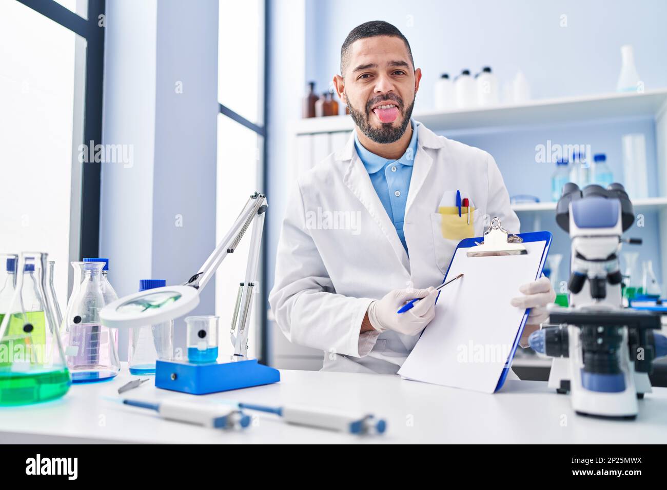 Hispanic man working at scientist laboratory holding blank clipboard sticking tongue out happy ...