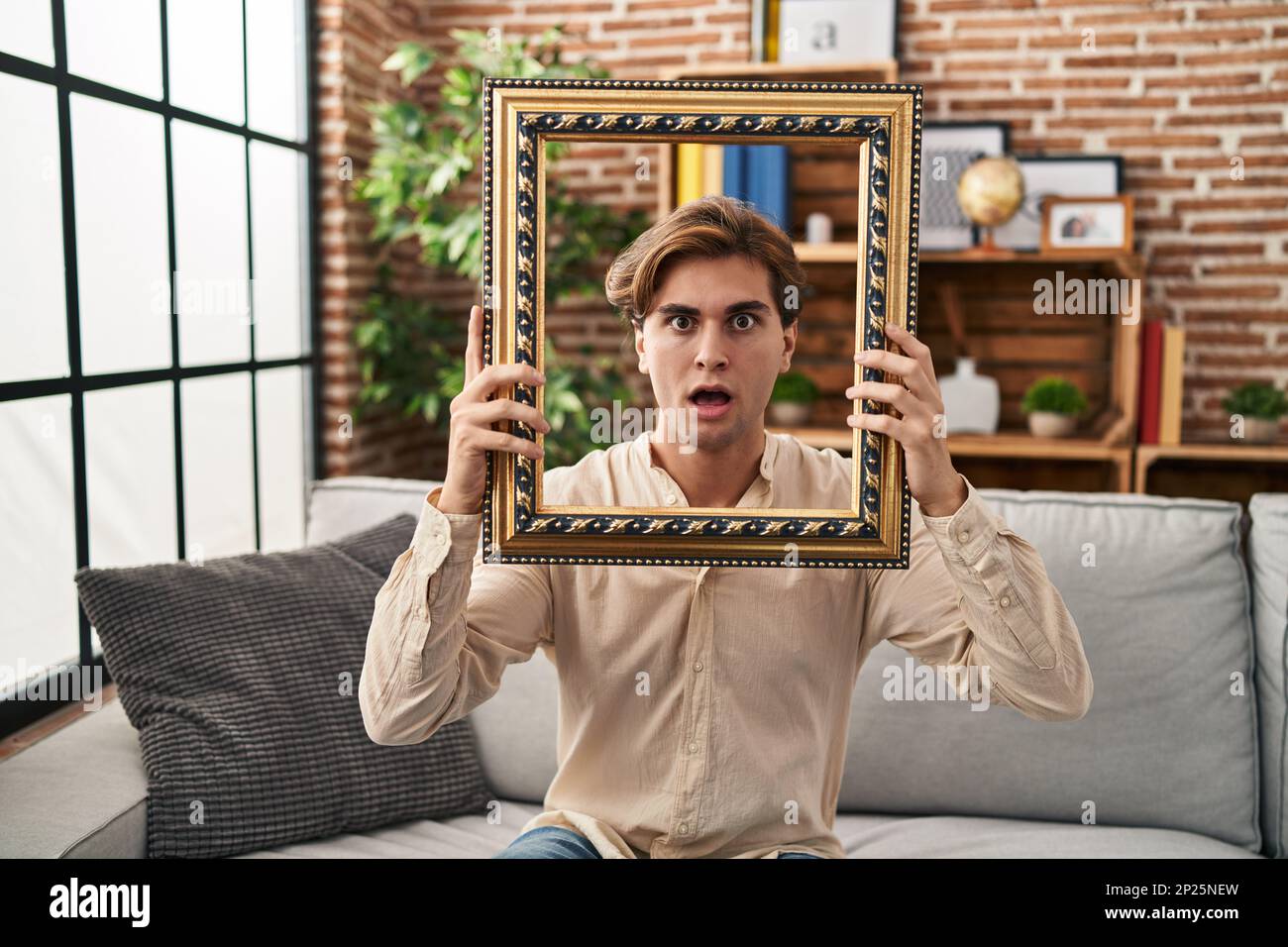 Young man holding empty frame in shock face, looking skeptical and ...