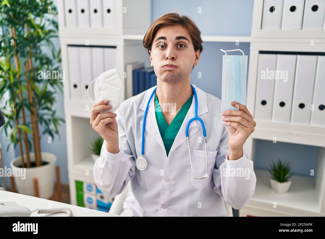 Young doctor man holding two safety mask puffing cheeks with funny face ...