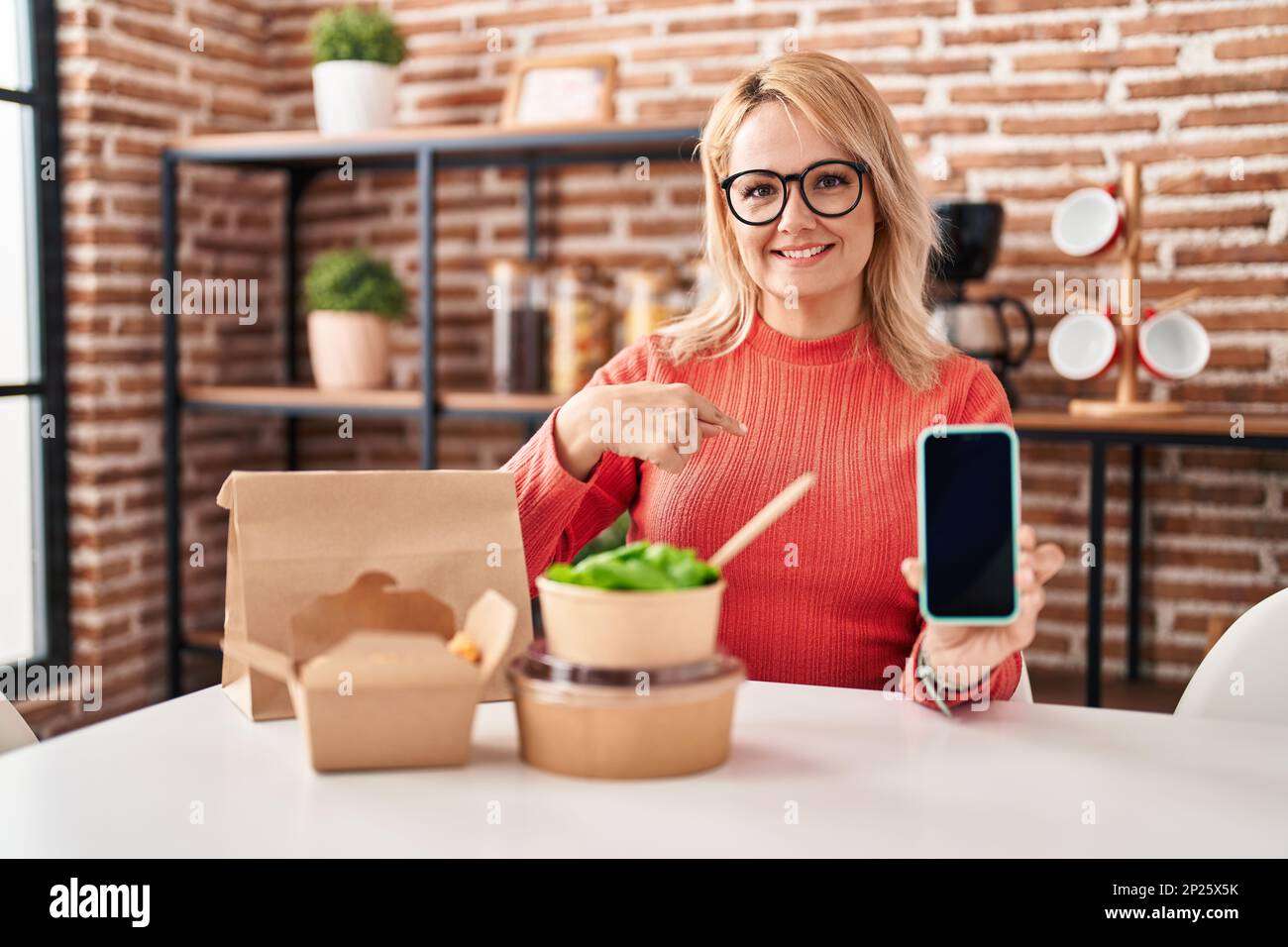 Blonde woman eating take away food showing smartphone screen pointing ...