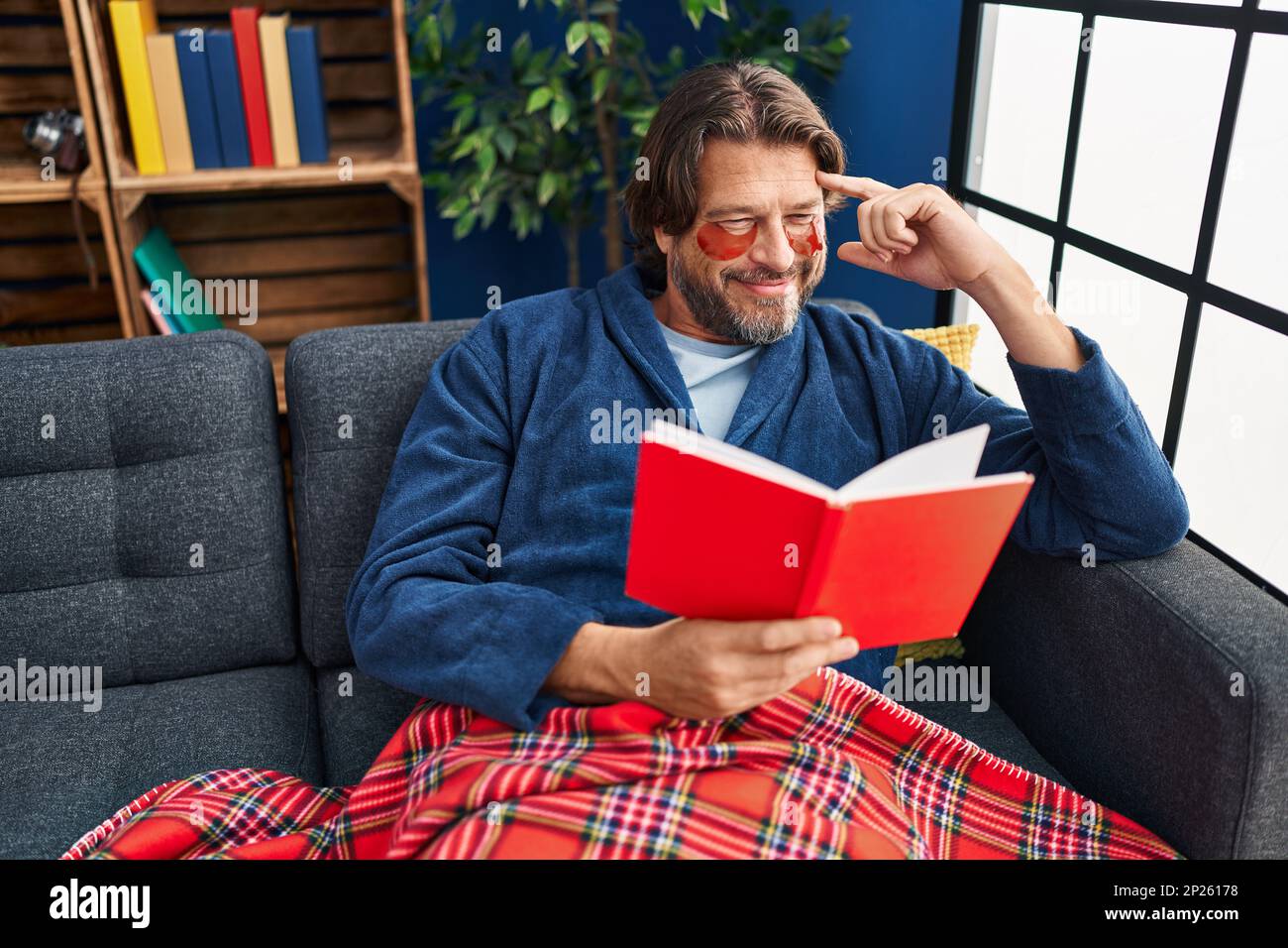 Middle age man wearing under eye patches reading book on sofa at home ...