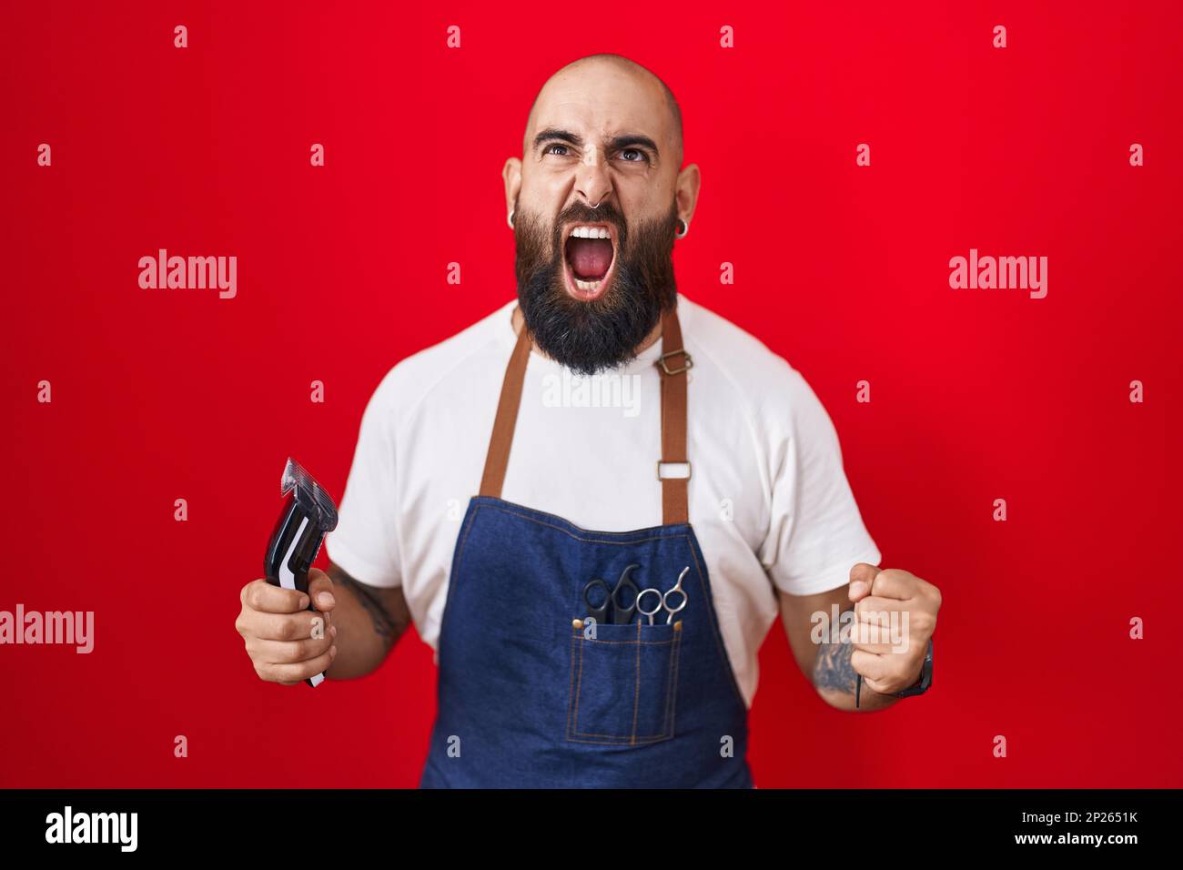 Young hispanic man with beard and tattoos wearing barber apron holding ...