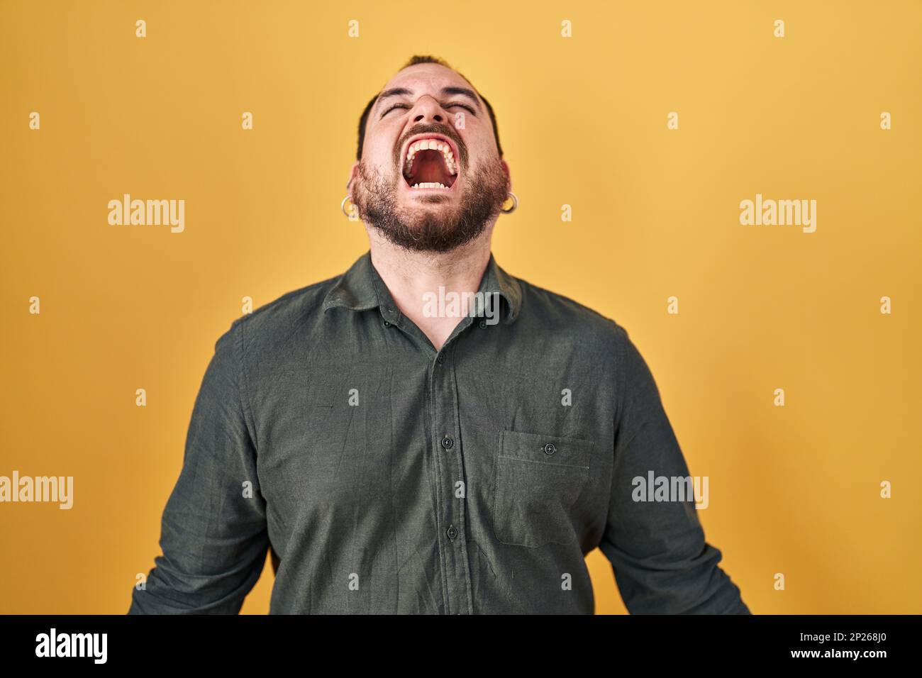 Plus size hispanic man with beard standing over yellow background angry ...